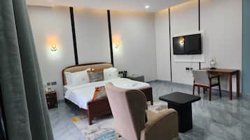 Quarto de hotel com cama, mesa de cabeceira, poltrona, escrivaninha, televisão e pequena mesa com abajur.
