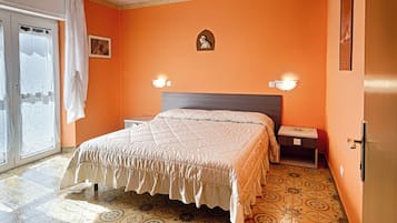 Quarto com cama, mesa de cabeceira, luminária na parede, porta e janela com cortinas.