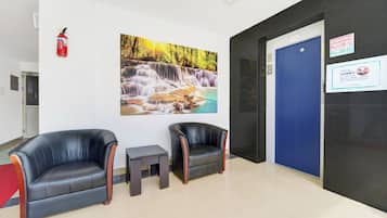 Área de recepção com duas poltronas de couro preto, uma mesa pequena, um extintor de incêndio, uma porta de elevador azul e uma placa fixada na parede.