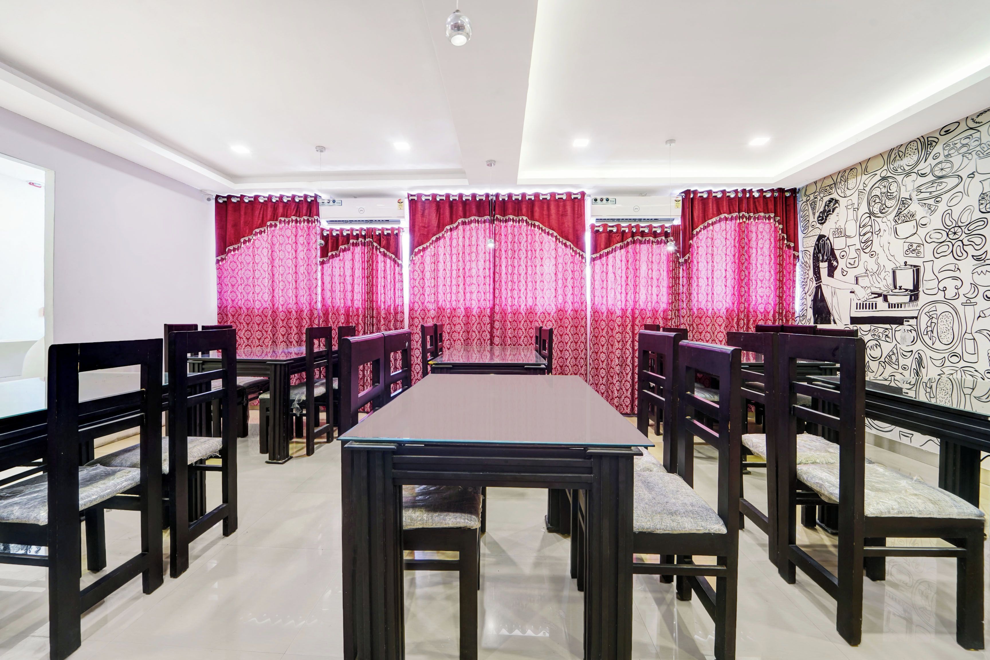 Uma sala de jantar com uma mesa comprida e cadeiras, cortinas vermelhas estampadas e uma parede com ilustrações de comida.