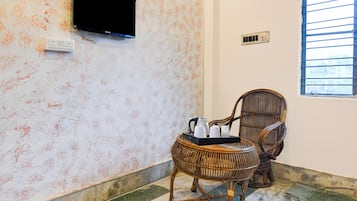Quarto com televisão na parede, uma cadeira de vime, uma mesa pequena com chaleira e xícaras, e uma janela com persianas.