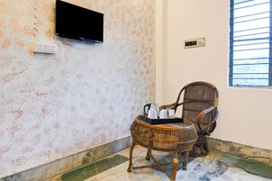 Quarto com televisão na parede, uma cadeira de vime, uma mesa pequena com chaleira e xícaras, e uma janela com persianas.