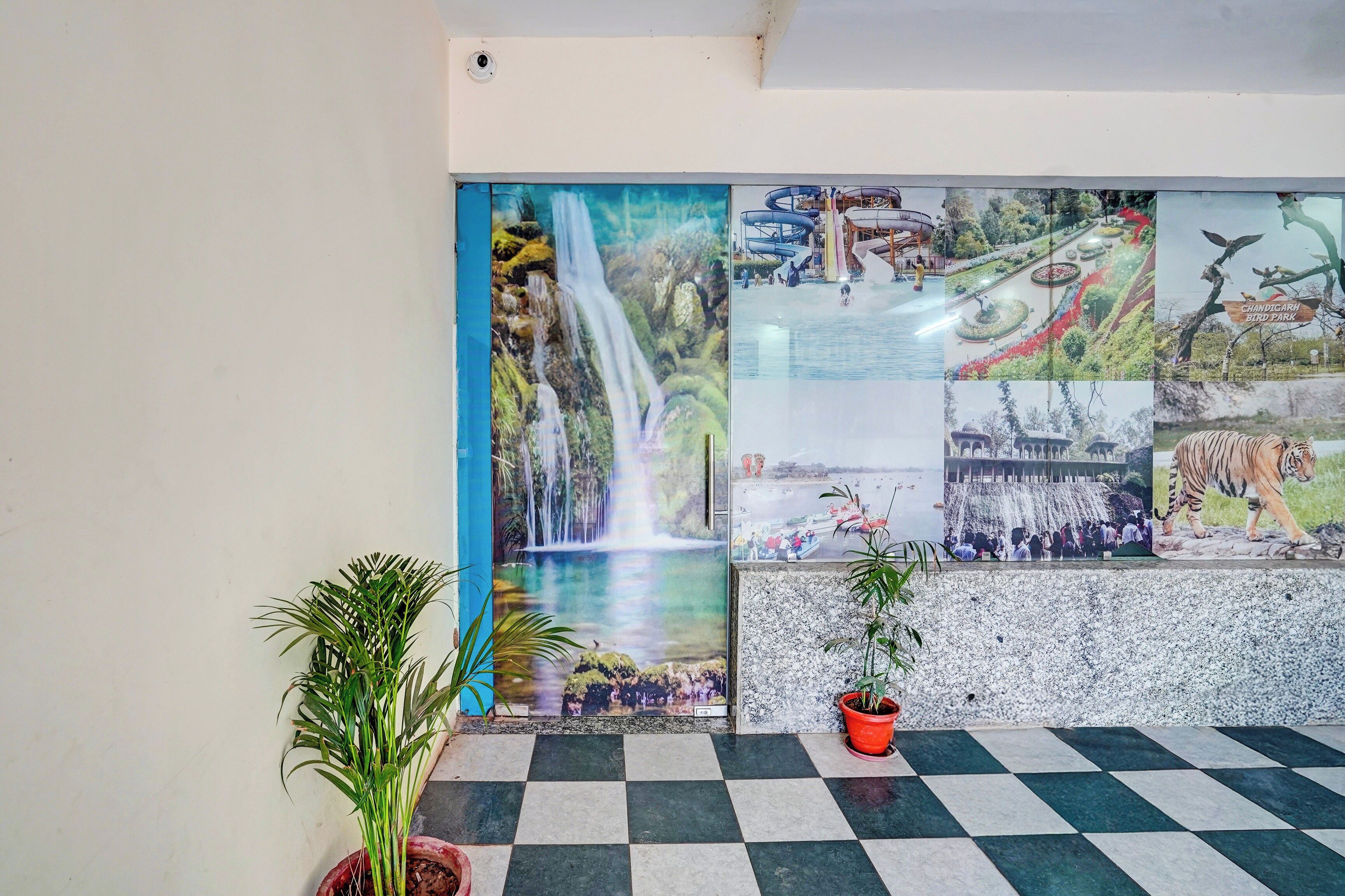 Uma área de recepção com piso xadrez, um mural grande com diversas cenas e duas plantas em vasos.