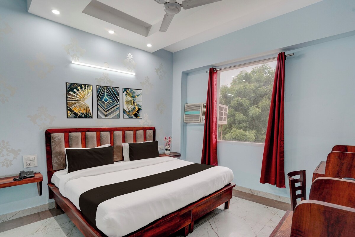Quarto de hotel com cama, mesa de cabeceira, telefone, janela com cortinas vermelhas e uma obra de arte na parede.