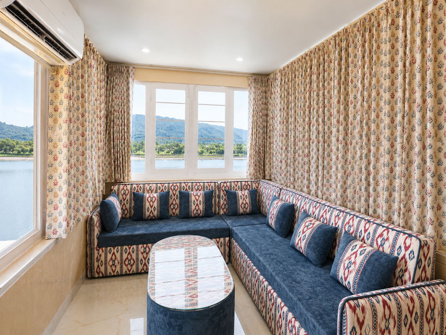 Um canto com área de estar, incluindo um sofá e uma mesa pequena, uma janela com cortinas e vista para um corpo d'água e colinas.