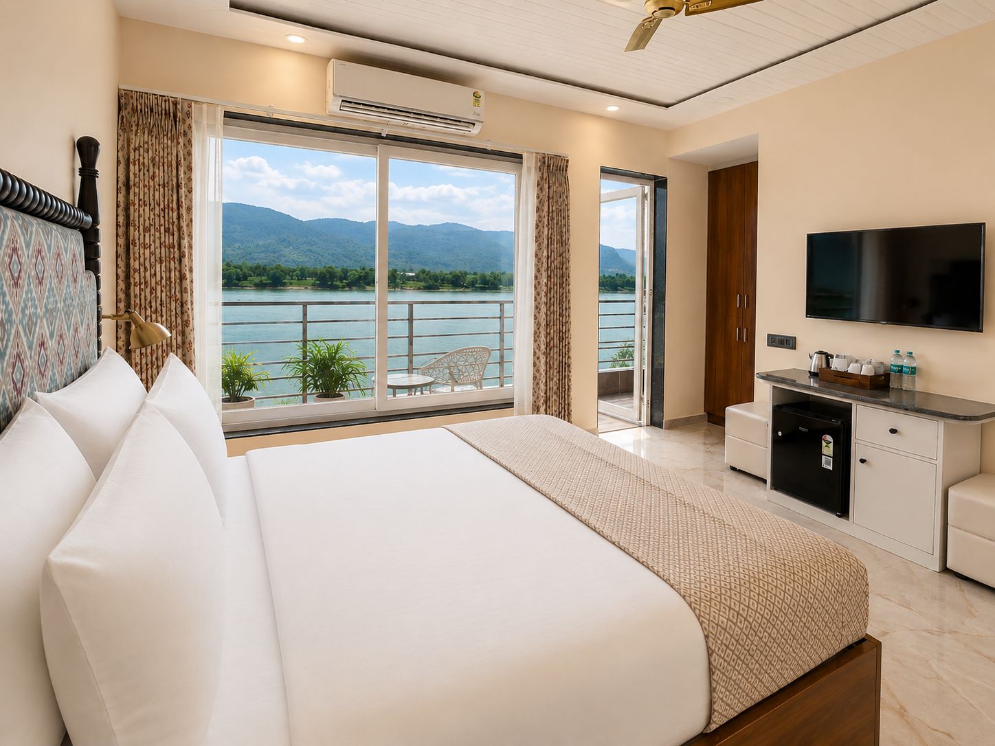 Quarto de hotel com uma cama grande, televisão, escrivaninha com cafeteira e vista para um lago e montanhas.