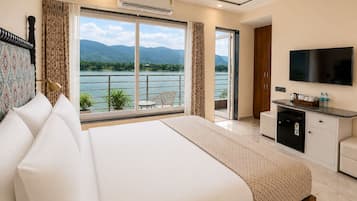 Quarto de hotel com uma cama grande, televisão, escrivaninha com cafeteira e vista para um lago e montanhas.