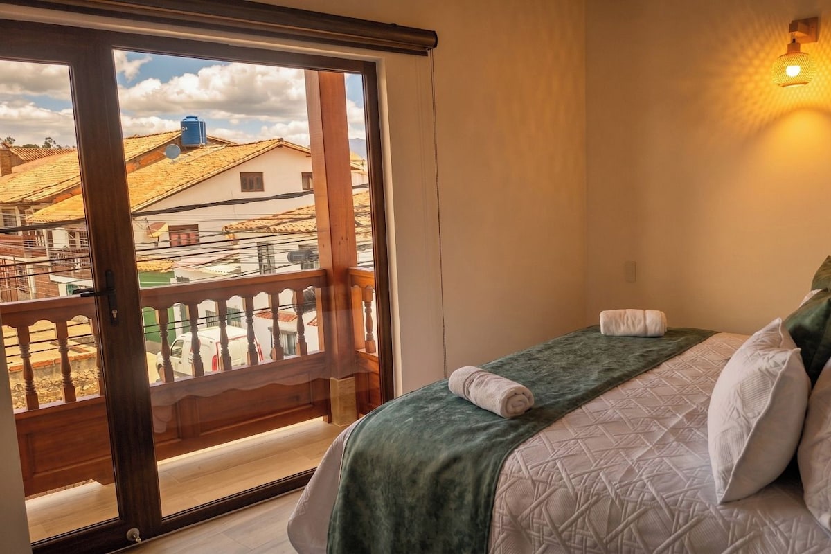 Quarto com cama, almofadas, manta verde e varanda com vista para edifícios e um reservatório de água azul.