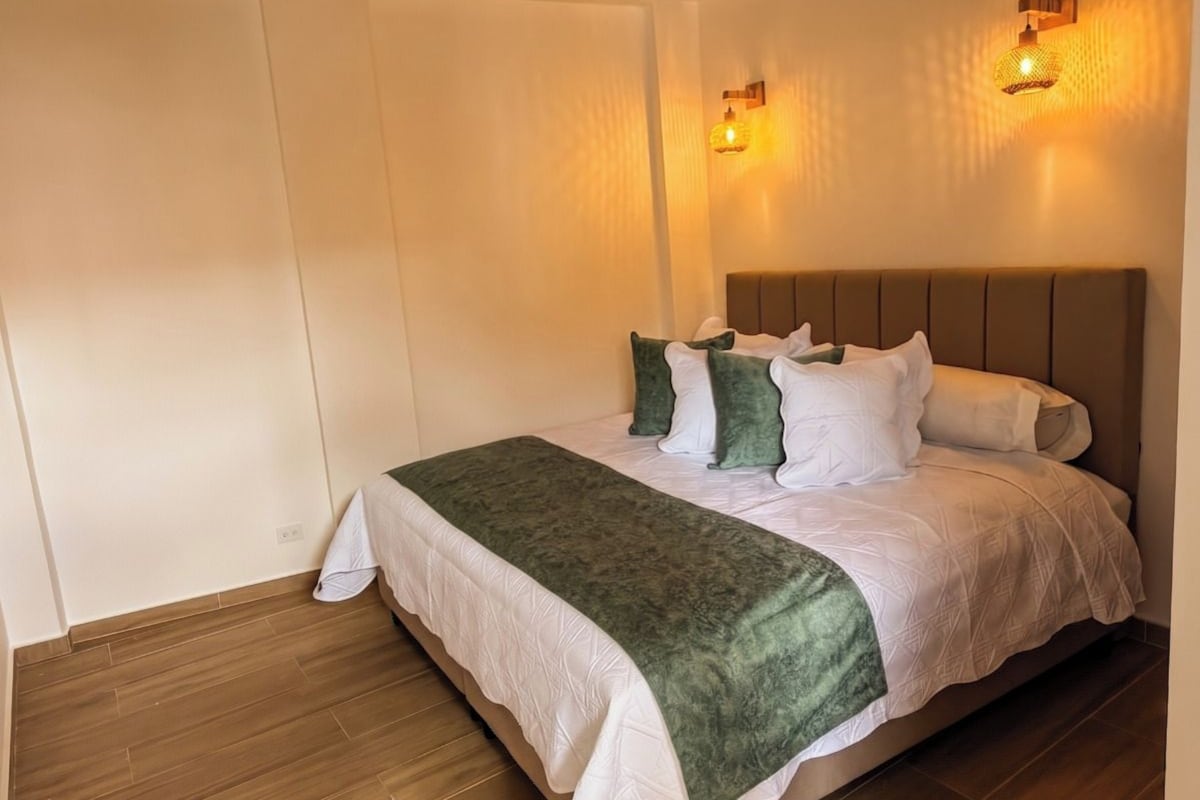 Quarto com cama, almofadas, cobertor, piso de madeira e iluminação fixa na parede.