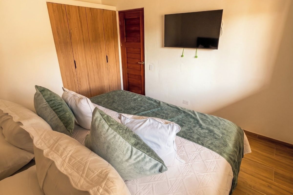 Quarto com cama, almofadas, guarda-roupa e televisão na parede.