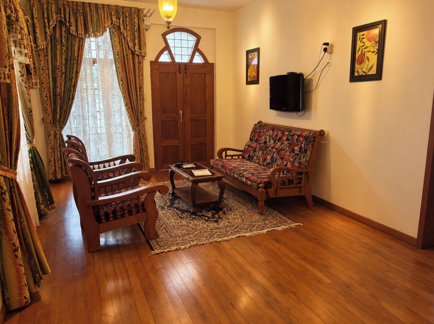 Uma sala de estar com sofá, cadeiras, mesa de centro, tapete, porta de madeira e TV fixada na parede.