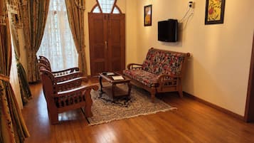 Uma sala de estar com sofá, cadeiras, mesa de centro, tapete, porta de madeira e TV fixada na parede.