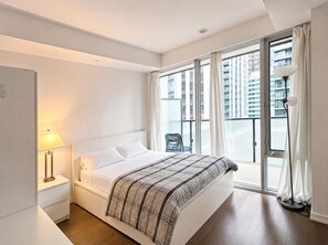 Quarto de hotel moderno com uma cama grande, uma mesa de cabeceira, uma luminária e vista da cidade por amplas janelas.