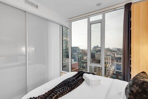Quarto de hotel com uma cama grande, vista para a paisagem urbana através de janelas do chão ao teto e uma porta deslizante.