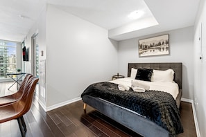 Quarto de hotel moderno com uma cama grande, vista da cidade e área de refeições com cadeiras.