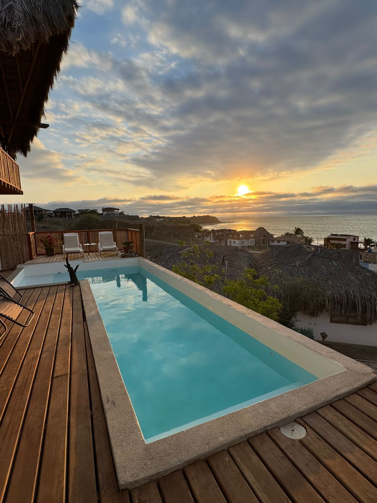 Piscina retangular com deck de madeira, rodeada por estruturas com telhado de palha, com vista para uma paisagem costeira com edifícios e um pôr do sol.
