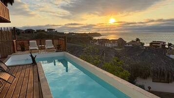 Piscina retangular com deck de madeira, rodeada por estruturas com telhado de palha, com vista para uma paisagem costeira com edifícios e um pôr do sol.