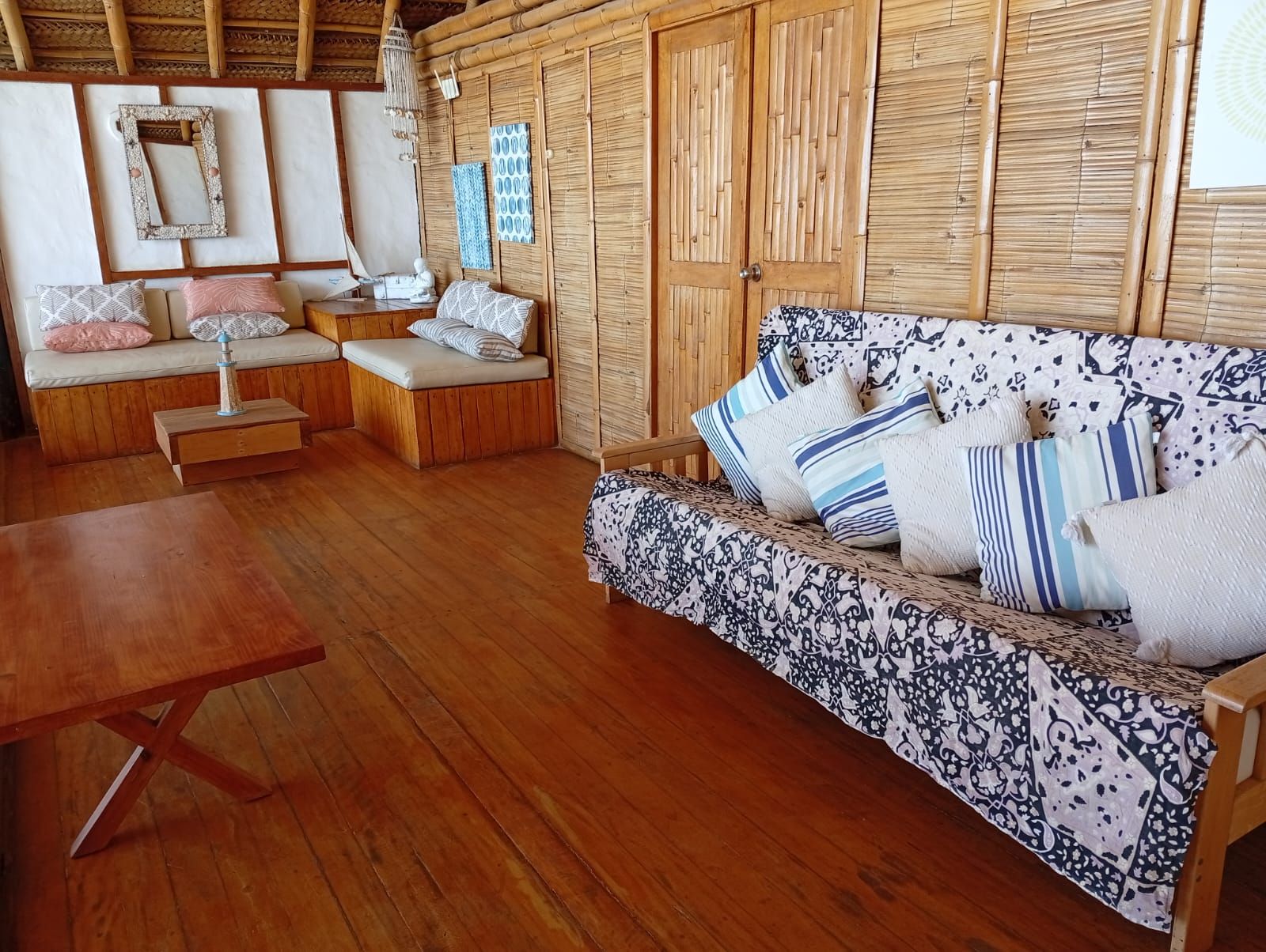 Quarto com piso de madeira, sofá com almofadas estampadas, mesa de madeira e parede com espelho e objetos decorativos.