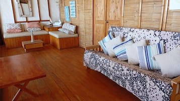Quarto com piso de madeira, sofá com almofadas estampadas, mesa de madeira e parede com espelho e objetos decorativos.
