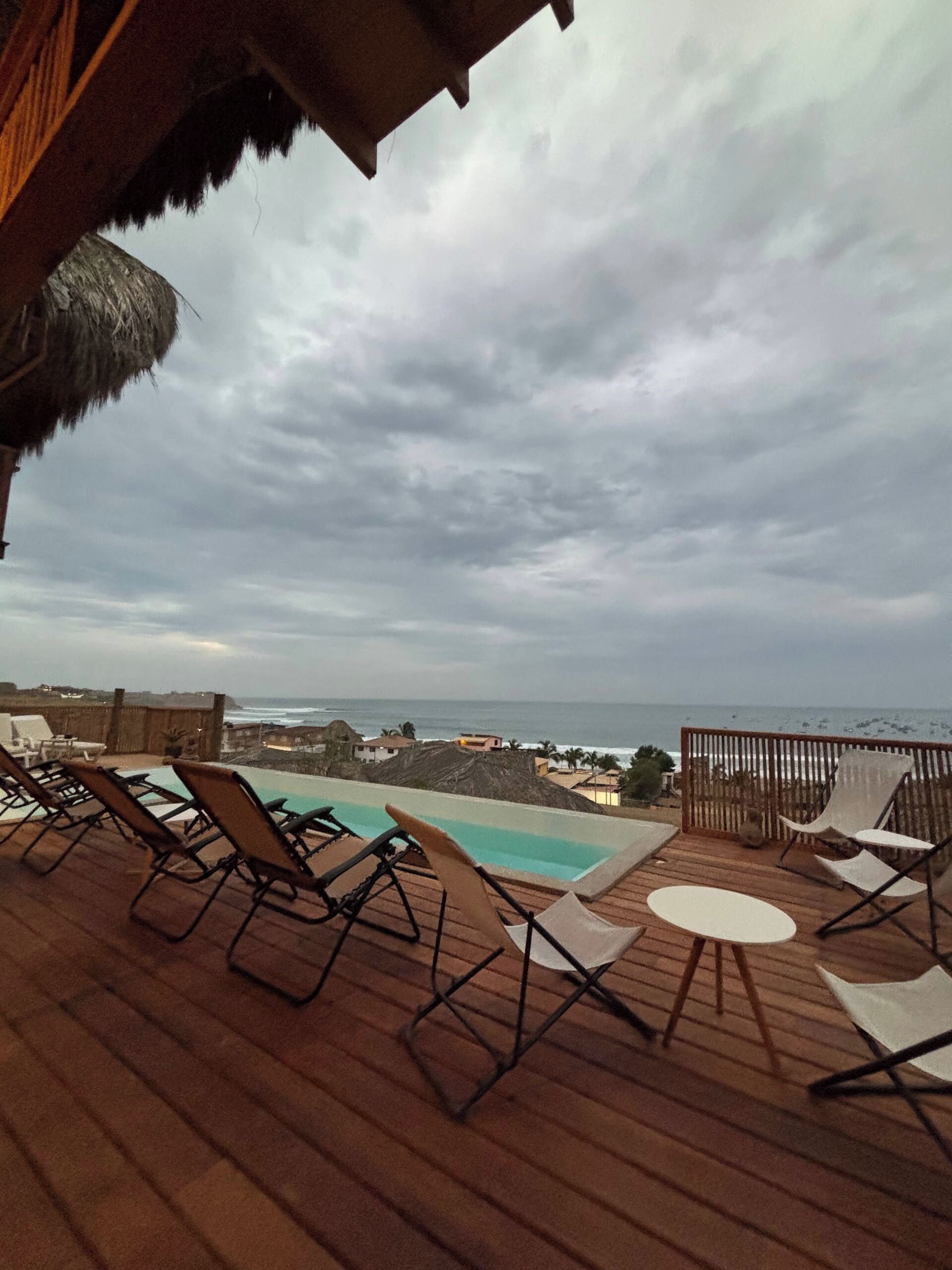 Um deck de madeira com cadeiras de descanso e uma piscina pequena, com vista para a área costeira, o oceano e edifícios ao longe.