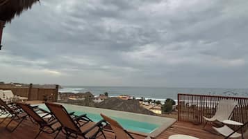 Um deck de madeira com cadeiras de descanso e uma piscina pequena, com vista para a área costeira, o oceano e edifícios ao longe.
