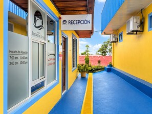 Uma área de recepção de hotel com balcão, janela com vista para o céu e placa indicando o nome do hotel e horário de funcionamento.