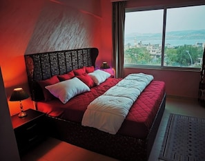 Quarto com uma cama grande, mesa de cabeceira, abajur e vista para a cidade pela janela.