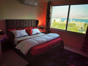 Quarto com uma cama grande, uma mesinha de cabeceira, uma luminária e vista para o horizonte urbano pela janela.