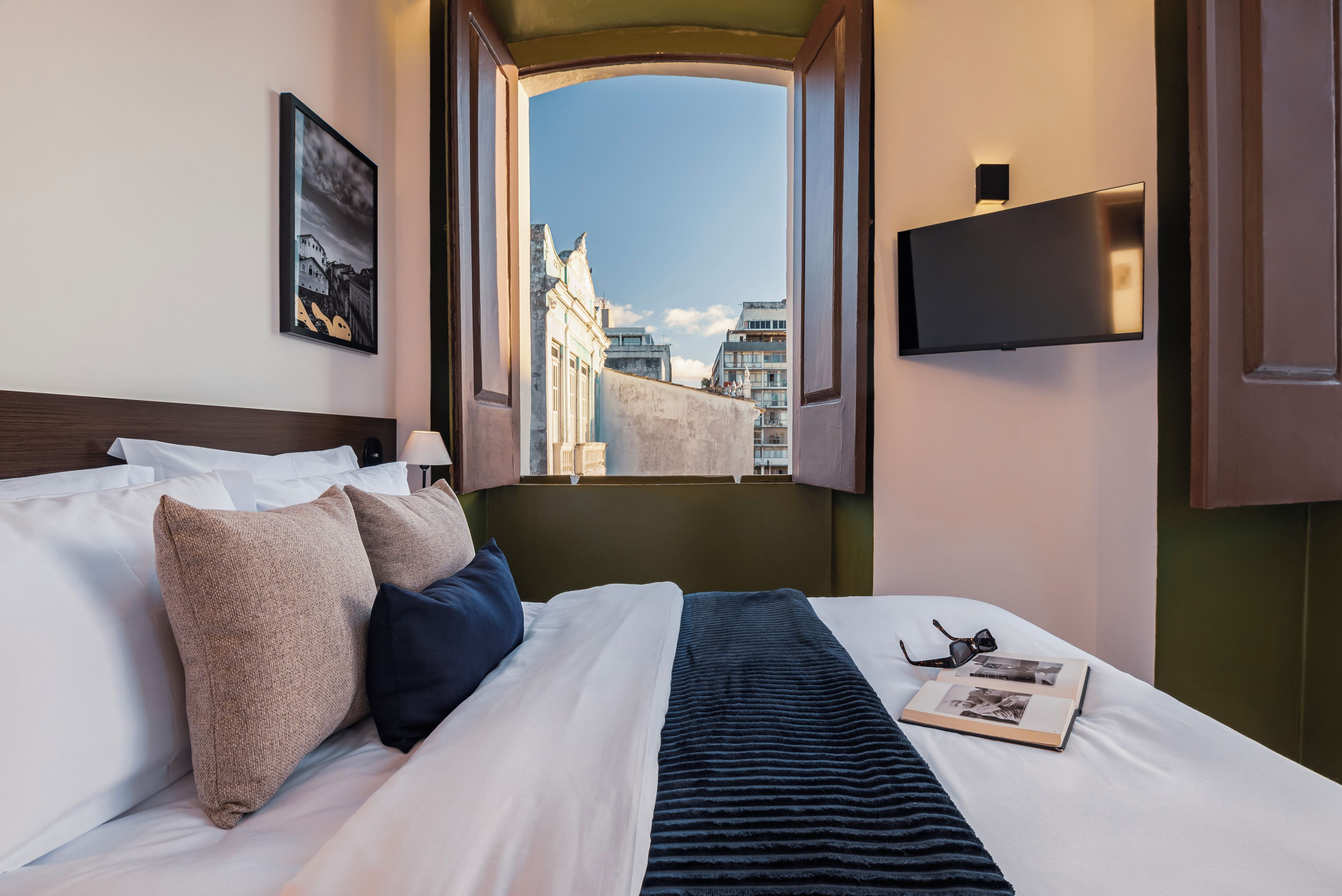 Quarto de hotel com cama, almofadas, mesa de cabeceira, livro, televisão e janela com vista para a cidade.