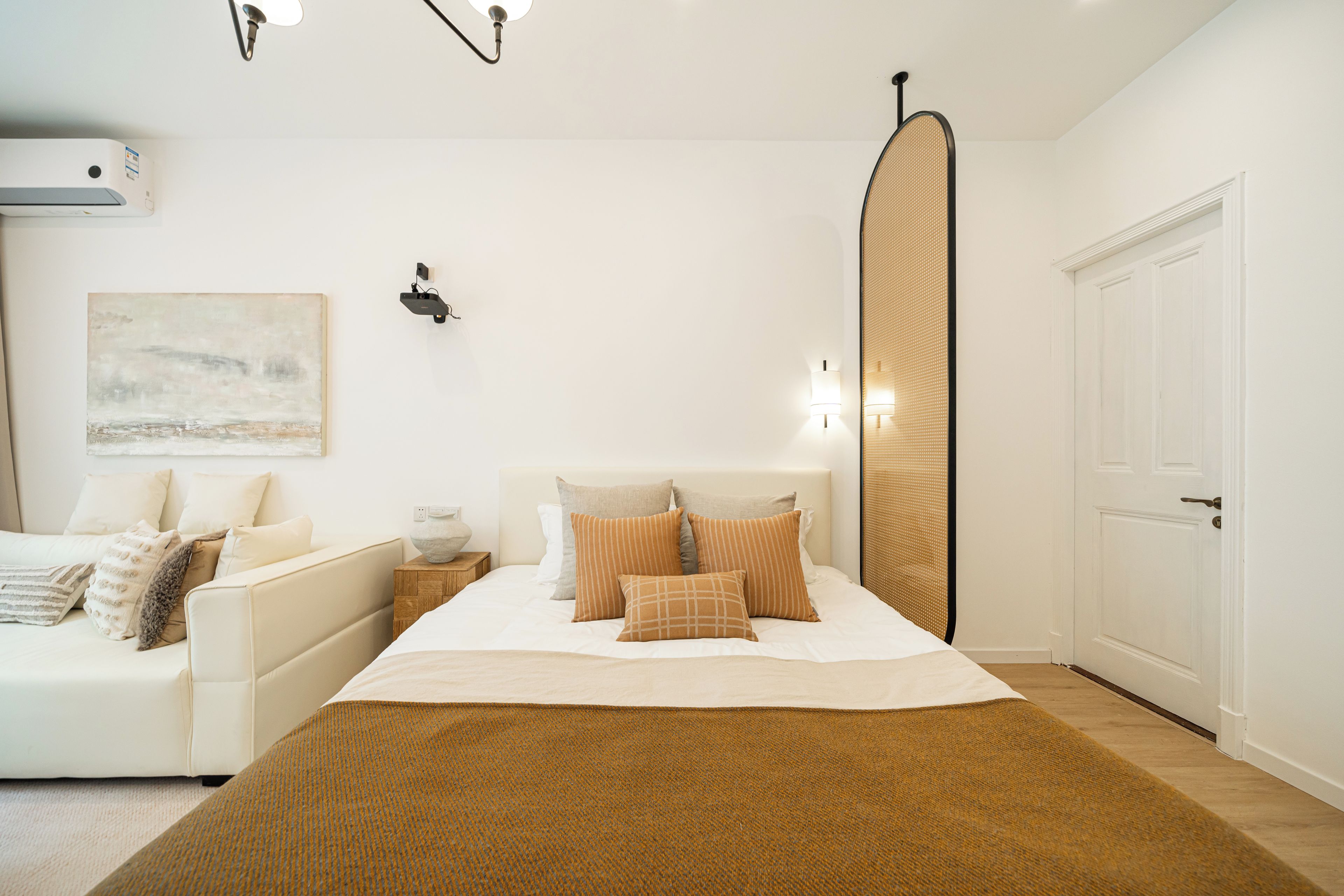 Quarto com cama, mesa de cabeceira, luminária na parede, um quadro e uma porta.
