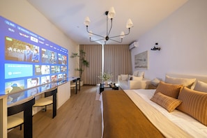Quarto de hotel moderno com uma tela grande exibindo opções de filmes, uma cama com várias almofadas, uma escrivaninha com cadeira, uma mesa de centro e um candelabro.