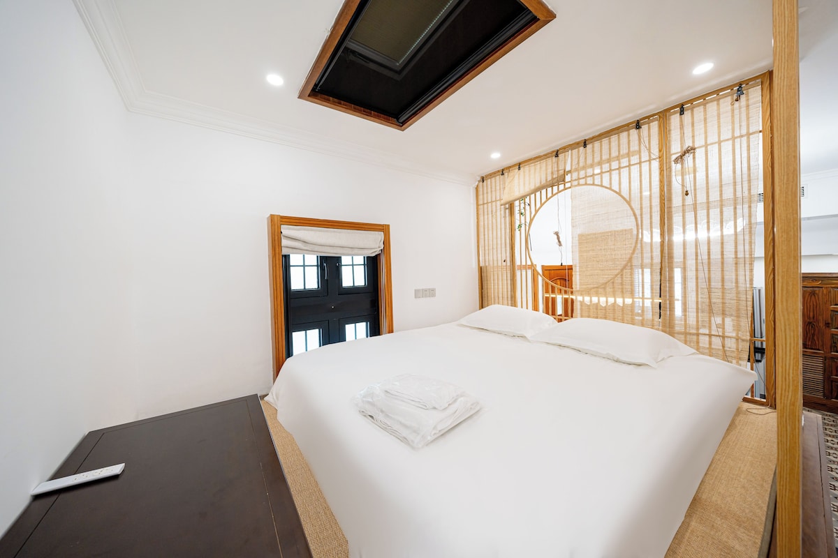 Quarto de hotel com cama, mesa de cabeceira, controle remoto, janela com persianas e luminária no teto.