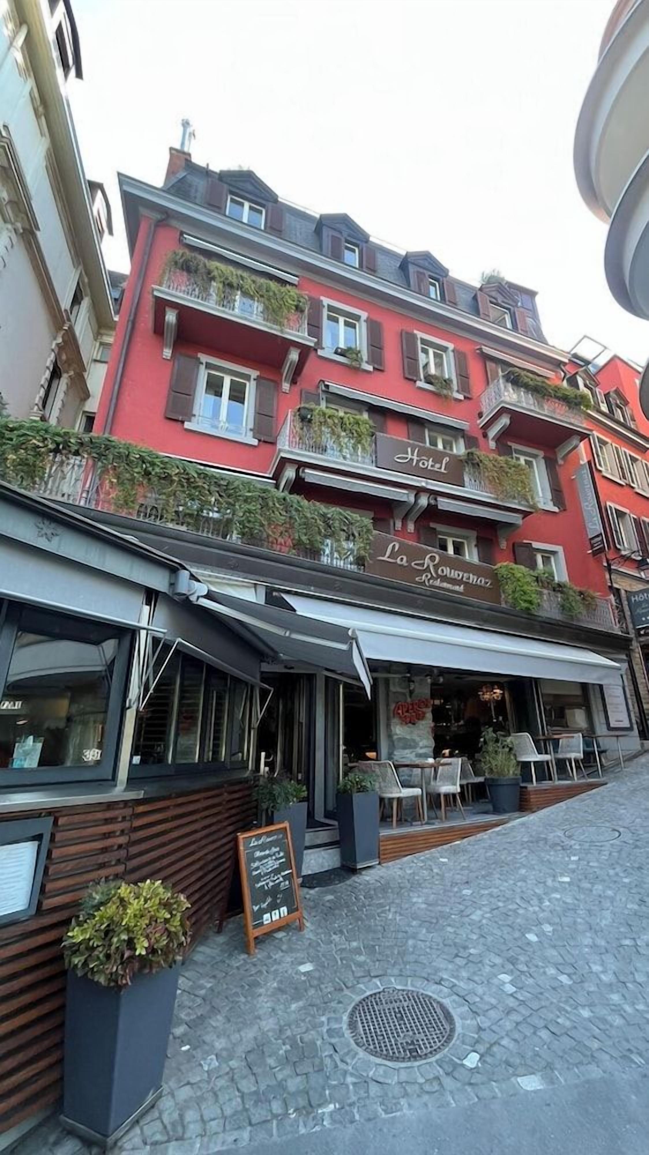 Um edifício vermelho com restaurante no andar térreo, placa com 'Hotel La Rouvière' e varanda com plantas.