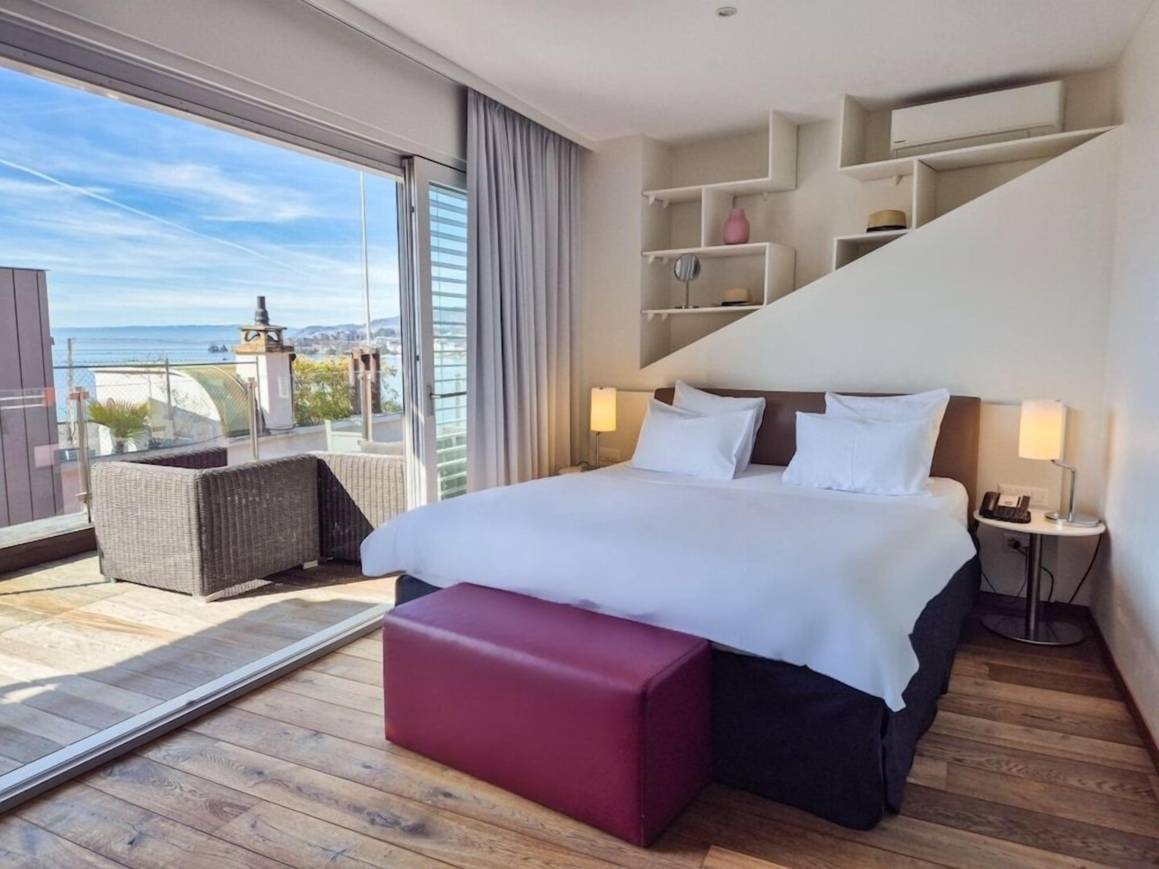 Quarto de hotel moderno com uma cama grande, um banco vermelho, varanda com vista e estantes embutidas.