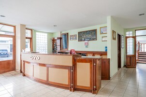 Área de recepção de hotel com balcão de madeira, uma janela grande e uma escadaria para outro andar.
