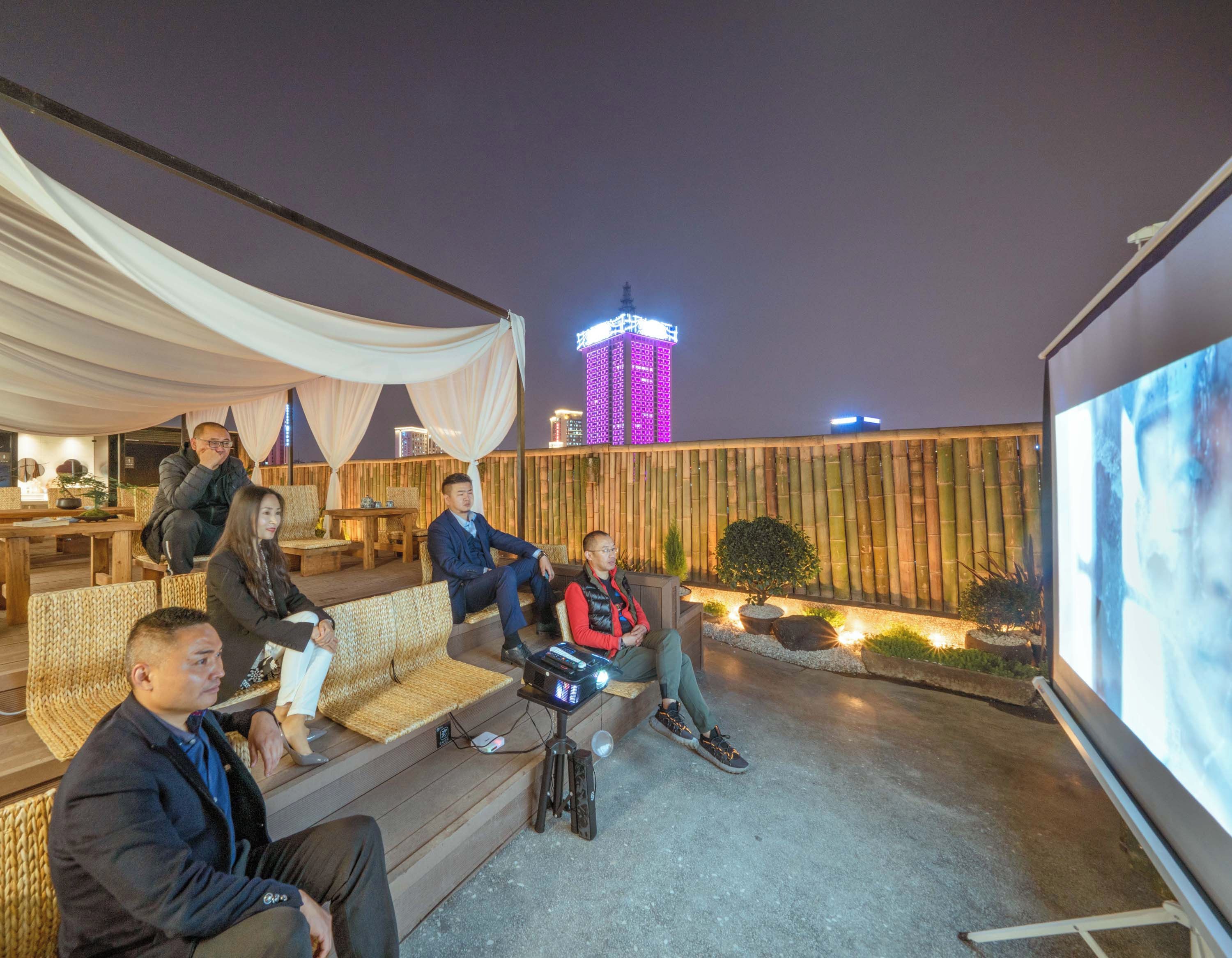 Um grupo de pessoas sentadas em móveis de exterior numa área coberta, com vista para a paisagem urbana, incluindo uma torre bem iluminada.