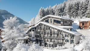 Um moderno edifício de estação de esqui com várias varandas e grandes janelas, rodeado por árvores e montanhas cobertas de neve.