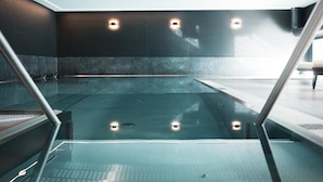 Piscina interna com piso de vidro, corrimãos de metal e iluminação no teto.