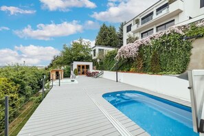 Uma área moderna com piscina ao ar livre, deck de madeira, um pequeno edifício e um prédio branco com jardim.