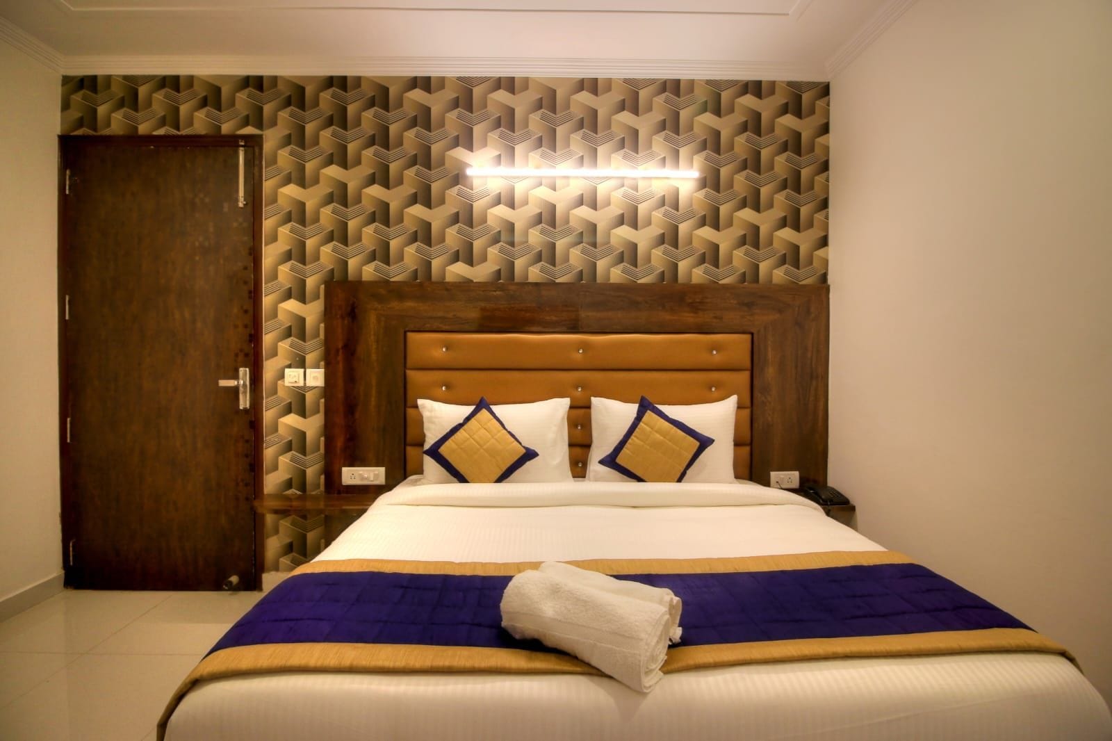 Quarto de hotel com cabeceira de madeira, papel de parede estampado, cama com colcha azul e dourada e toalha branca sobre a cama.