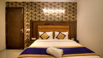 Quarto de hotel com cabeceira de madeira, papel de parede estampado, cama com colcha azul e dourada e toalha branca sobre a cama.
