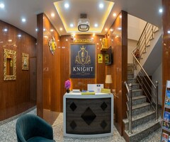 Uma área de recepção com balcão, uma placa com a inscrição 'The Knight Guest House' e uma escadaria à direita.