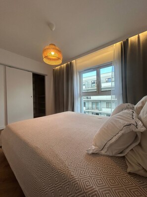 Quarto com cama, janela com cortinas, luminária no teto e vista para um edifício.