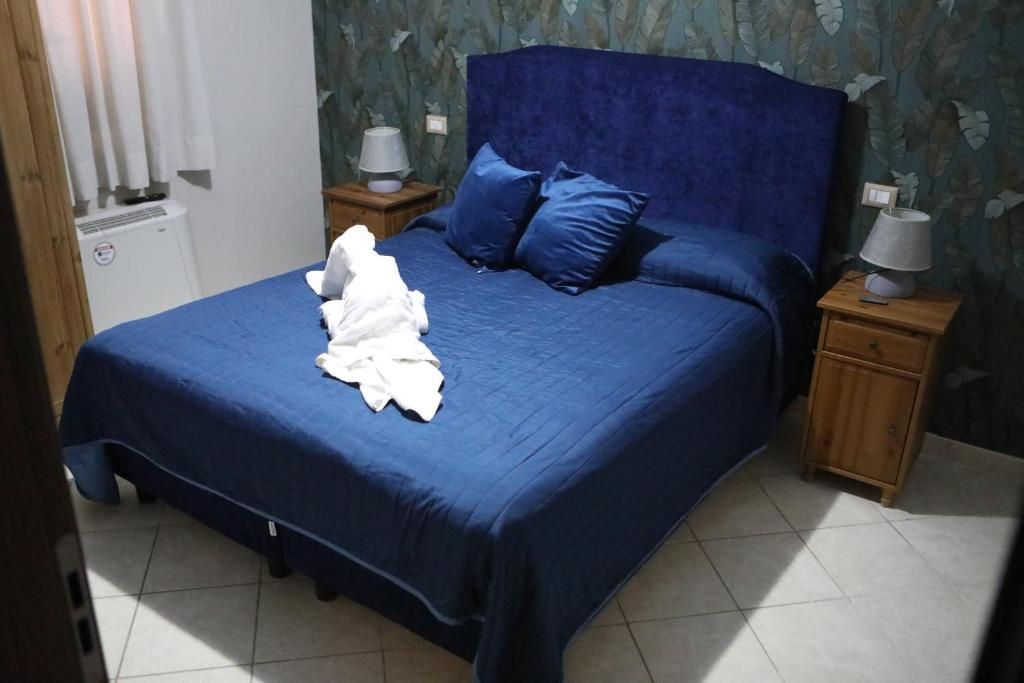 Cama com cabeceira azul e travesseiros, mesa de cabeceira com abajur e radiador na parede.