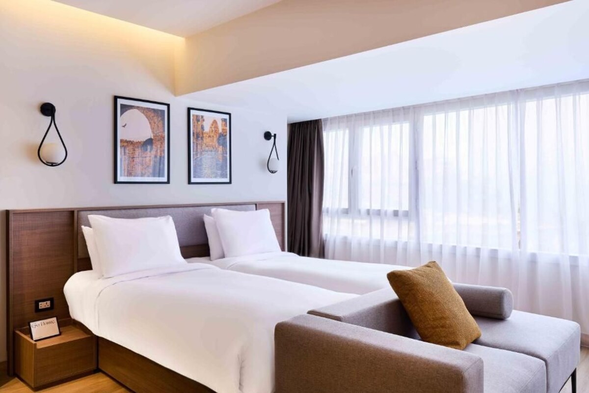 Quarto de hotel com duas camas, mesa de cabeceira, sofá e janela com cortinas.