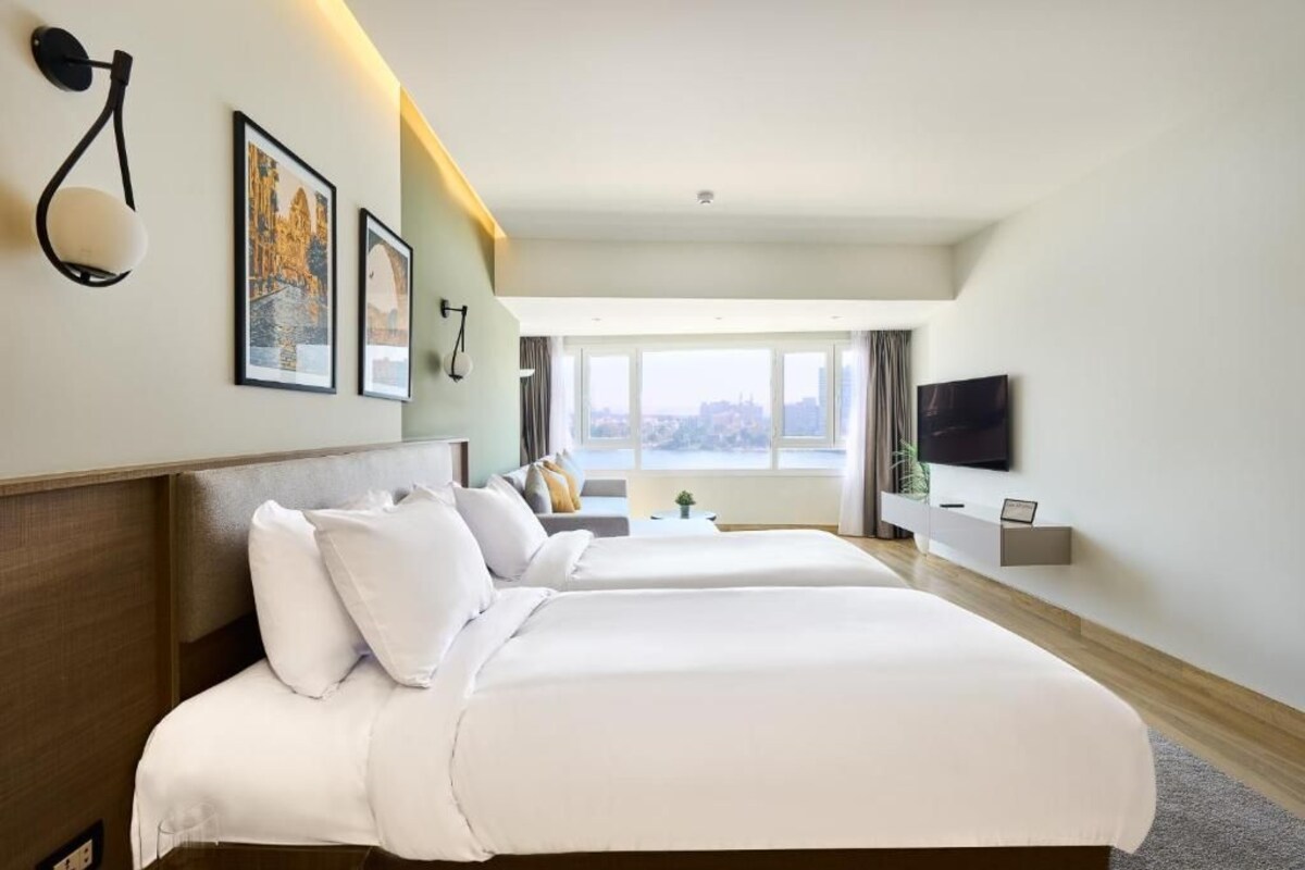 Quarto de hotel com duas camas, televisão, janela com vista para a cidade e obras de arte na parede.
