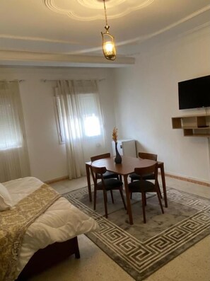 Quarto de hotel com uma cama, uma mesa de jantar com cadeiras, uma pequena TV na parede e um tapete estampado.