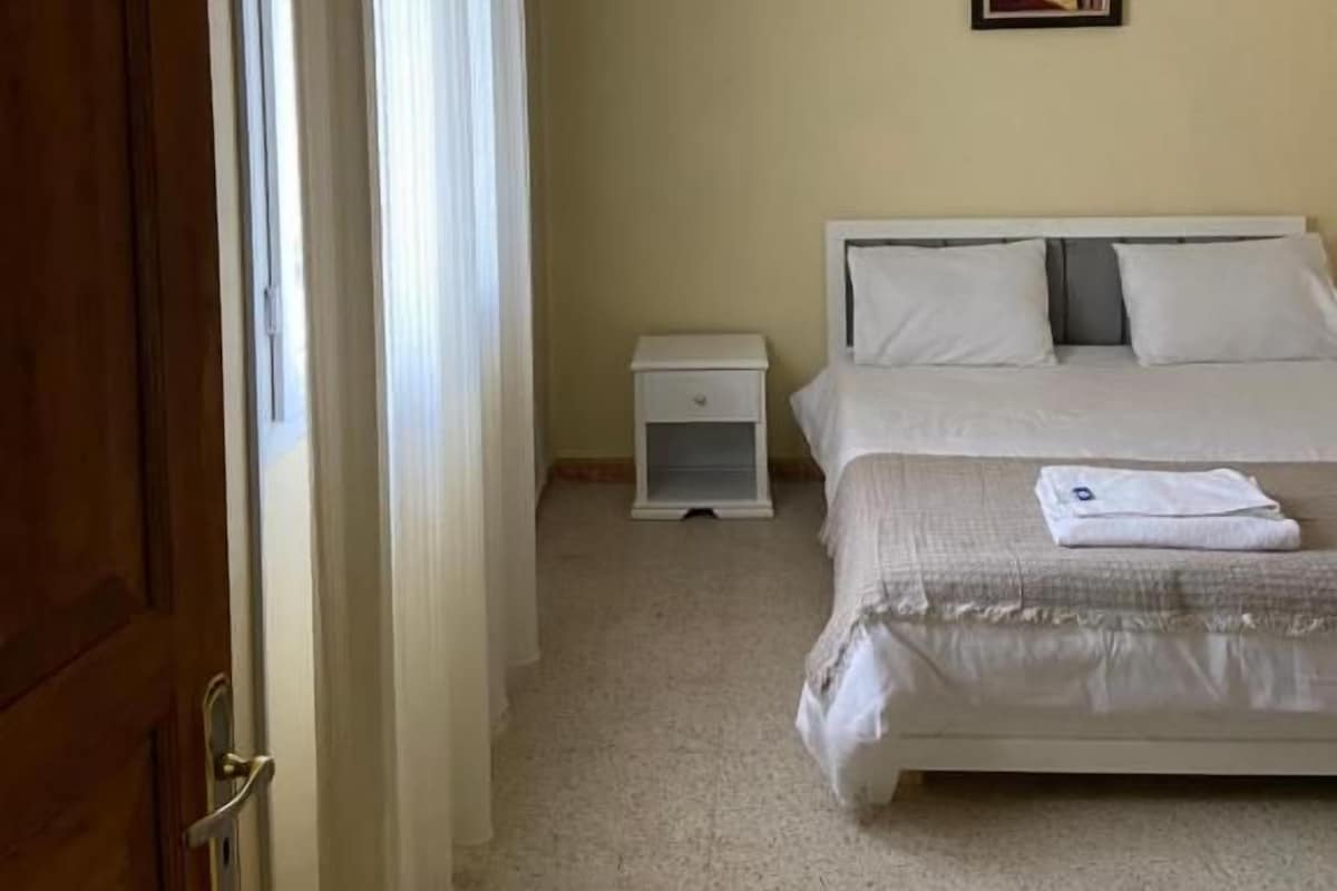 Quarto com cama, mesa de cabeceira, porta e ventilador de teto.