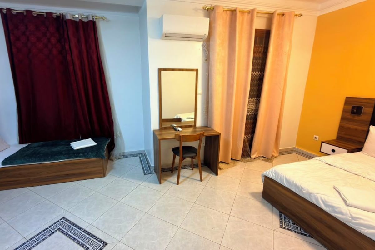 Quarto de hotel com cama, escrivaninha e cadeira, espelho e janela com cortinas.