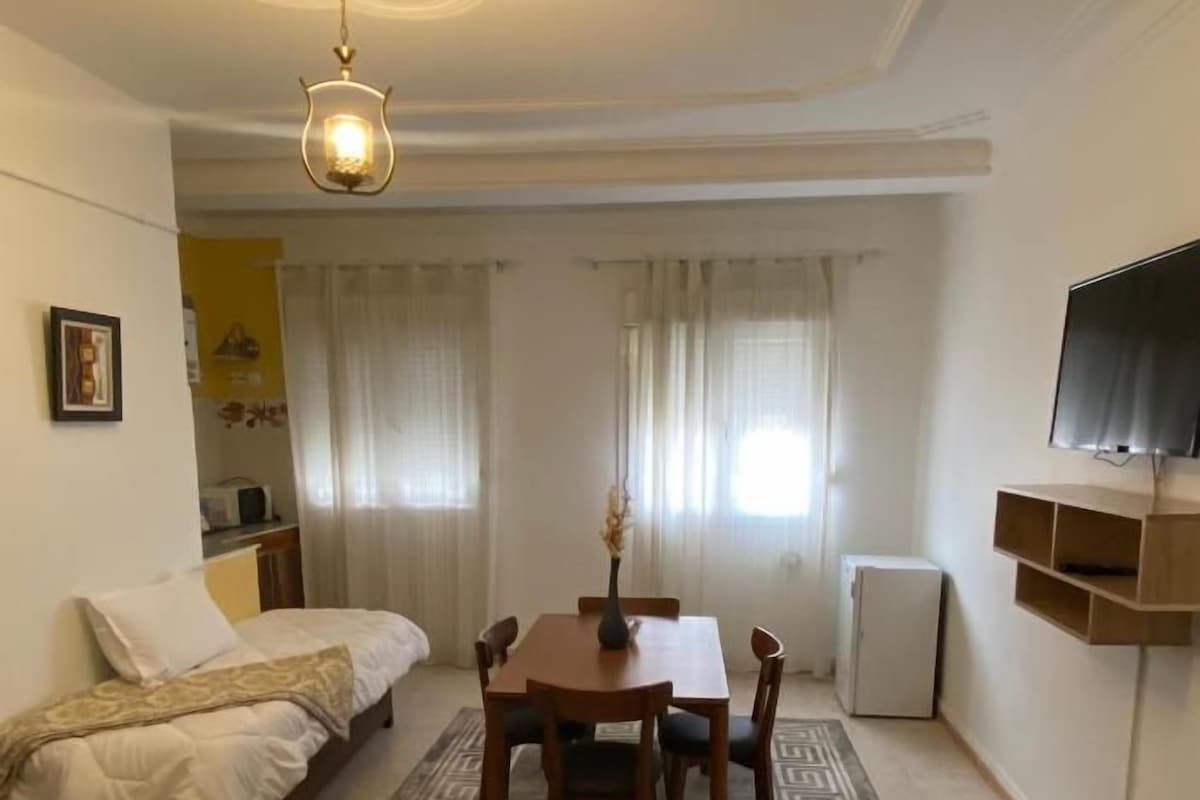 Quarto compacto com cama, mesa de jantar com cadeiras, uma TV pequena e um tapete decorativo.
